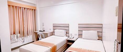 Quarto de hotel com duas camas de solteiro, uma escrivaninha, uma cadeira, uma mesa pequena e uma janela com cortinas.