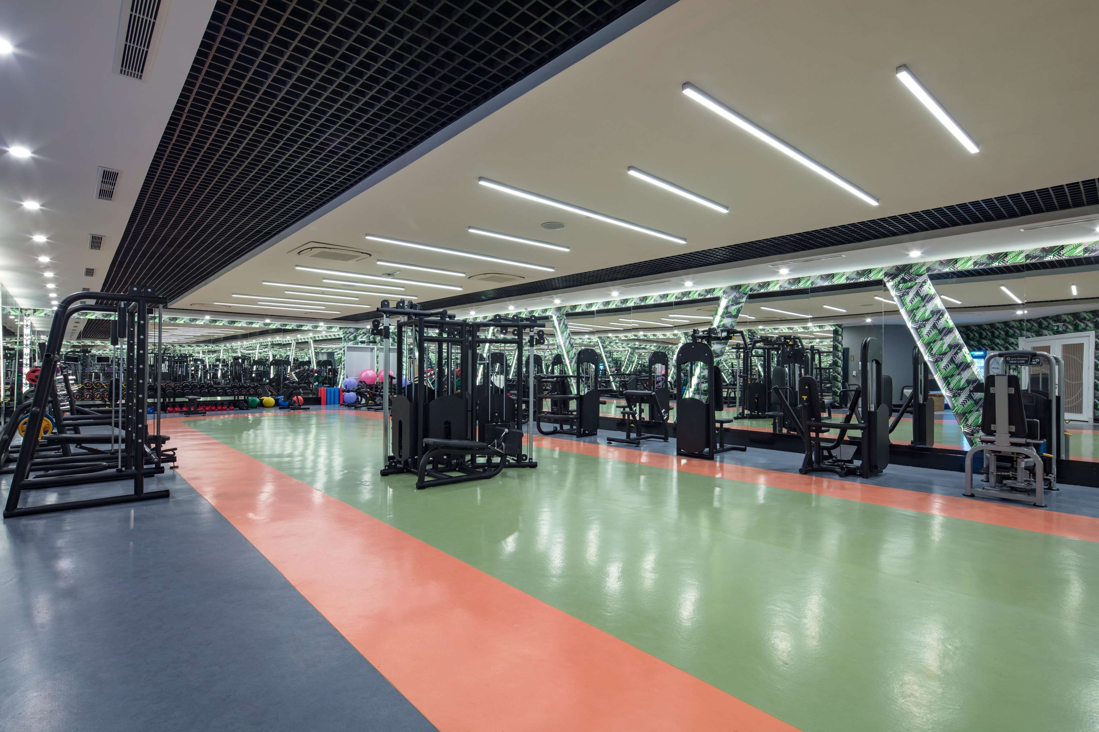 Uma academia espaçosa com diversos equipamentos de exercício, piso verde e laranja e um espelho grande na parede.