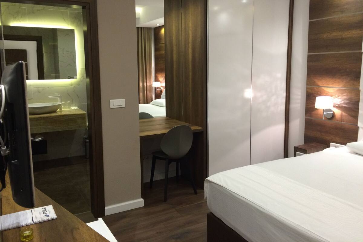 Quarto de hotel com cama, escrivaninha, cadeira, espelho, pia e guarda-roupa.