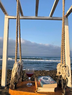 Uma cadeira de balanço de madeira com cordas, um copo de vinho tinto e um livro sobre um deck de madeira à beira-mar.