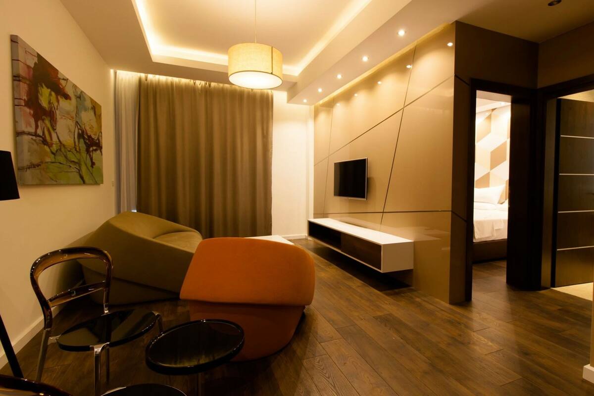 Quarto de hotel moderno com sofá, cadeiras, televisão, cama e uma janela grande com cortinas.