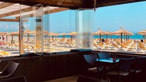 Restaurante à beira-mar com grandes janelas para uma praia, com espreguiçadeiras e guarda-sóis.