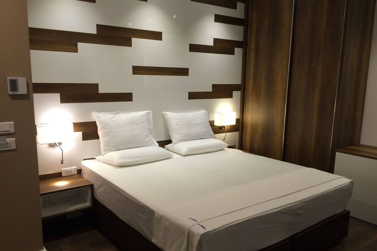 Cama bem arrumada com lençóis brancos e duas almofadas, abajur e parede com padrão geométrico.