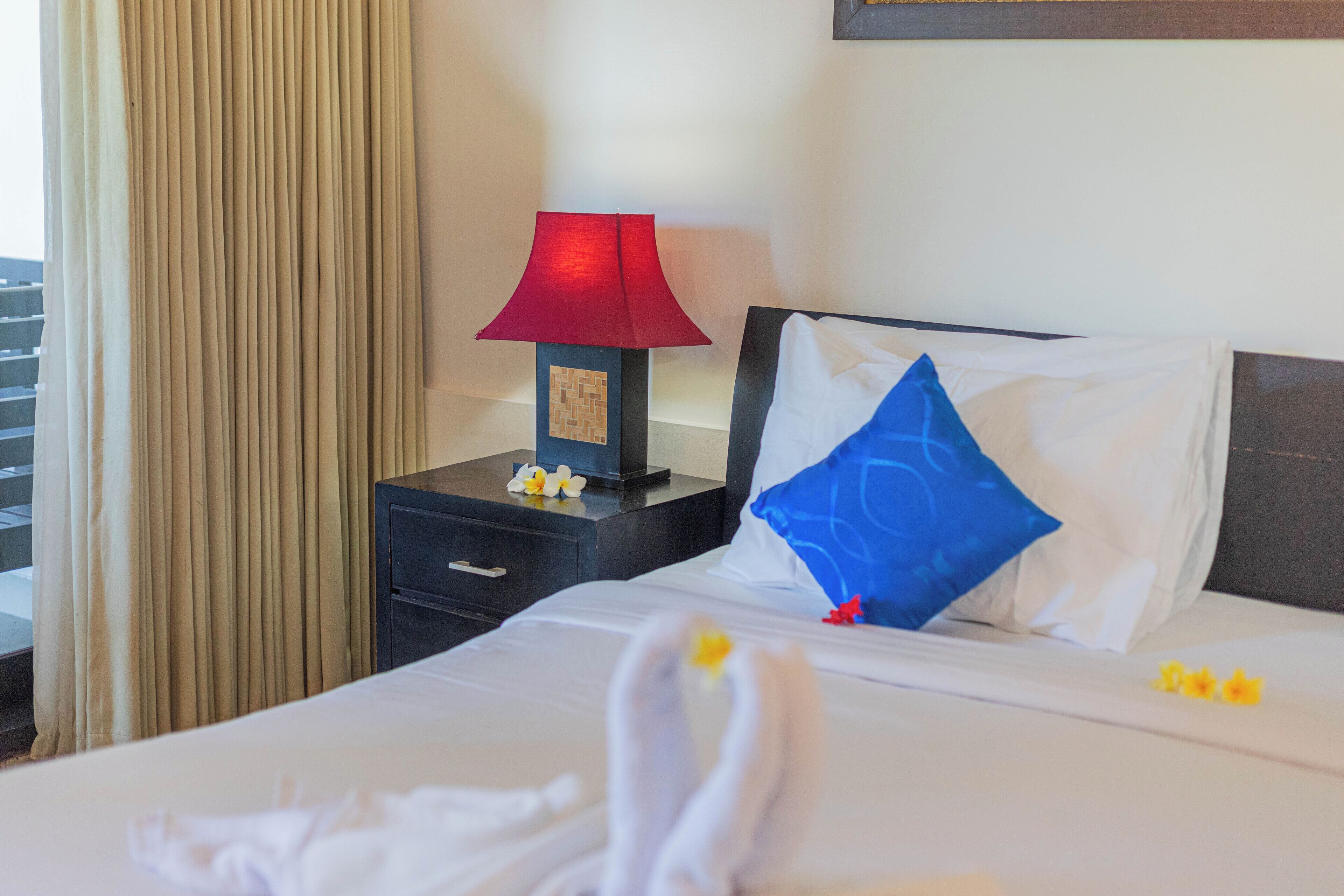 Quarto de hotel com cama, mesa de cabeceira com abajur vermelho e vista para o exterior.