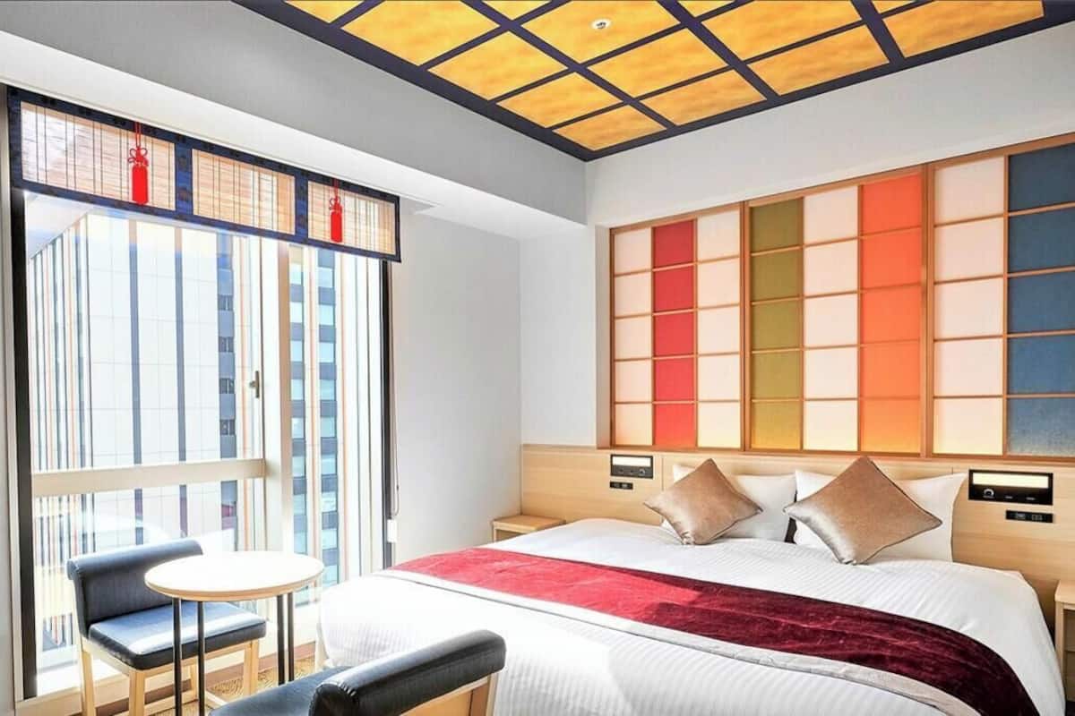 Quarto de hotel com cama, uma mesinha, uma cadeira, uma janela grande e um painel de parede em estilo tradicional japonês.