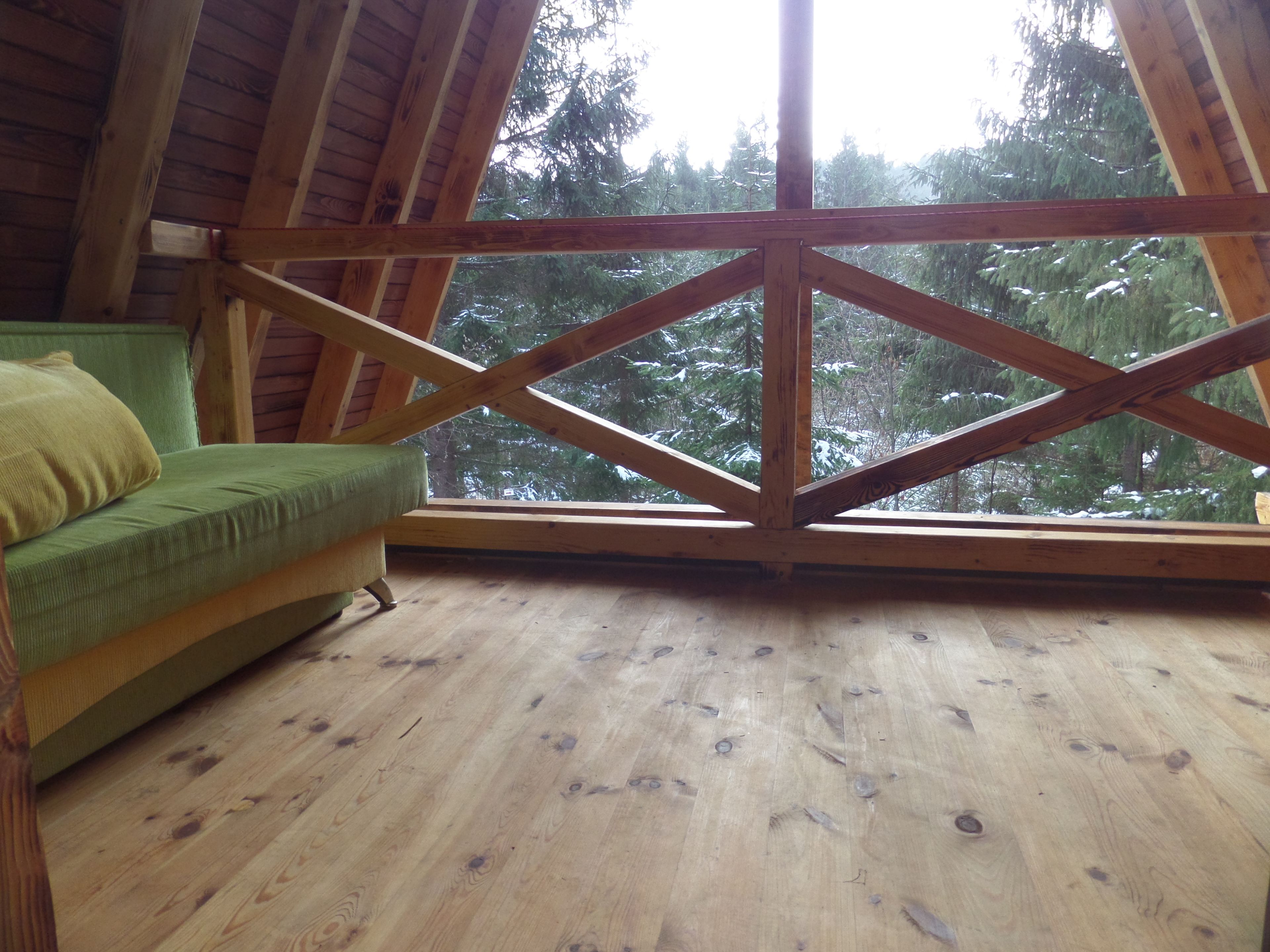 Quarto de cabana de madeira com sofá verde, janelas amplas e estrutura de teto em madeira.