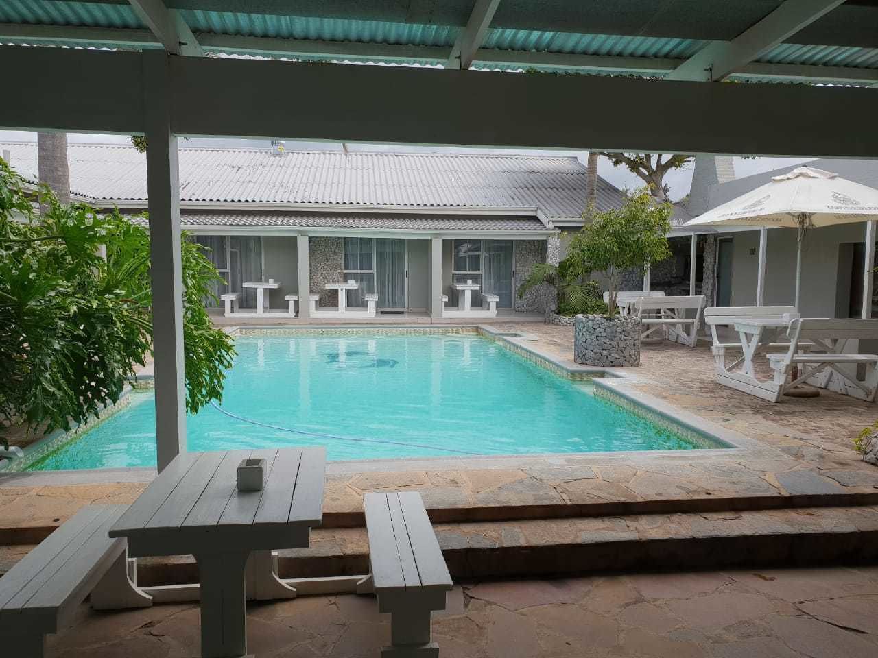 Uma área de piscina com um alpendre coberto, assentos externos e um edifício ao fundo.