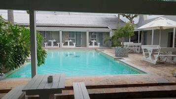Uma área de piscina com um alpendre coberto, assentos externos e um edifício ao fundo.