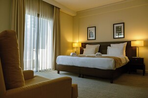 Quarto de hotel com uma cama grande, duas mesas de cabeceira com abajures, uma cadeira e uma janela ampla com cortinas.