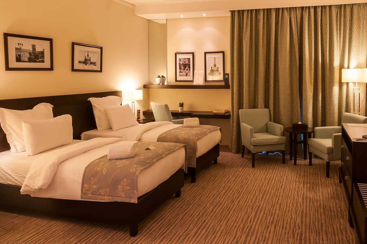 Quarto de hotel com duas camas, uma escrivaninha, cadeiras e quadros na parede.