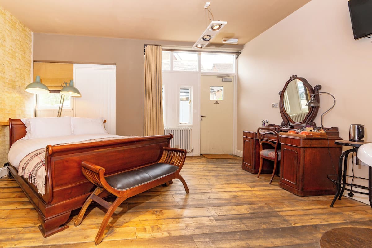 Quarto com cama de madeira, um banco, escrivaninha, cadeira, espelho e radiador.