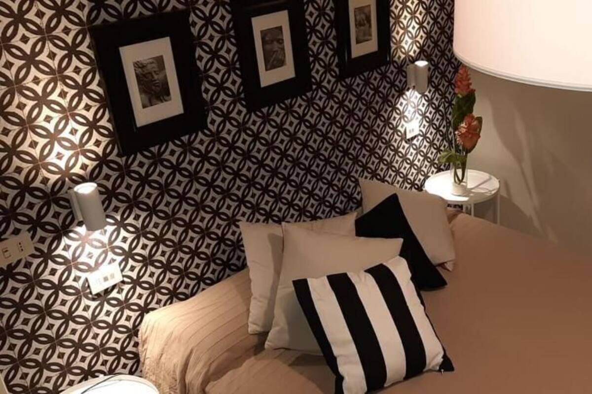 Quarto de hotel com papel de parede estampado, cama com almofadas listradas, mesa de cabeceira com abajur e um vaso com flores.