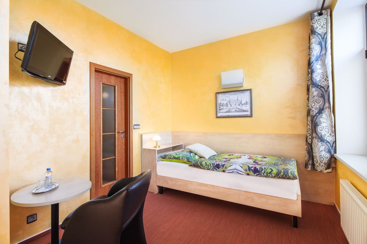Quarto pequeno com uma cama de solteiro, cabeceira de madeira, mesa de cabeceira com abajur, televisão de tela plana na parede, mesinha redonda com uma cadeira e janela com cortina estampada.