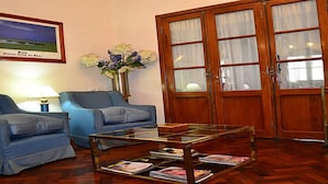 Quarto com dois sofás azuis, uma mesa de centro de vidro, portas duplas de madeira, um quadro na parede e um vaso com flores.