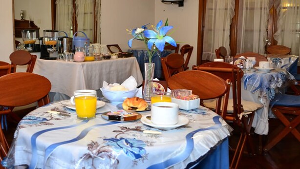 Uma sala de jantar com mesas redondas cobertas por toalhas de mesa florais, cadeiras de madeira e itens de café da manhã como croissants, café e suco.