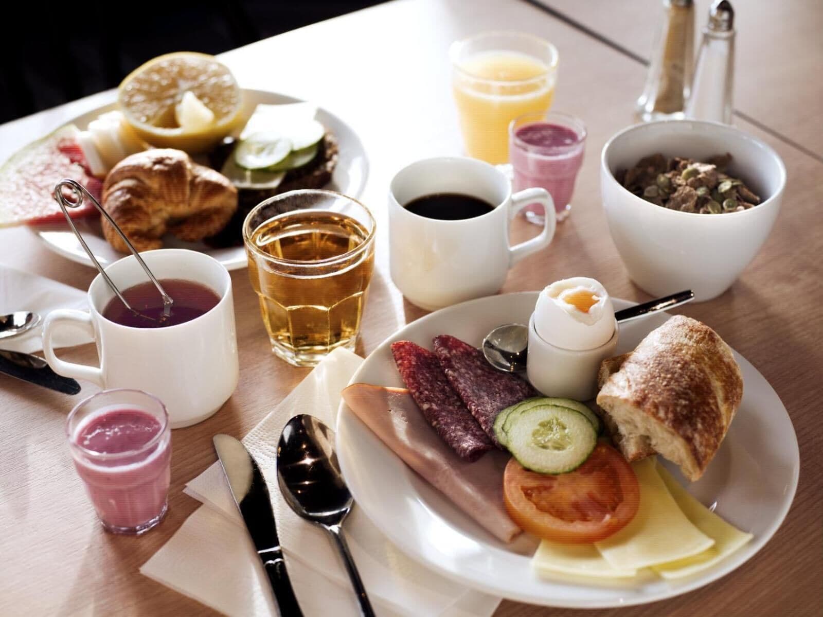 Disposição para o café da manhã com pratos de comida, bebidas e utensílios sobre uma mesa.