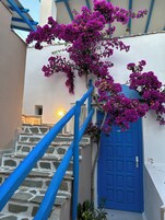 Uma escadaria com corrimãos azuis que leva a uma porta azul, emoldurada por paredes brancas e flores de bougainvillea roxas.