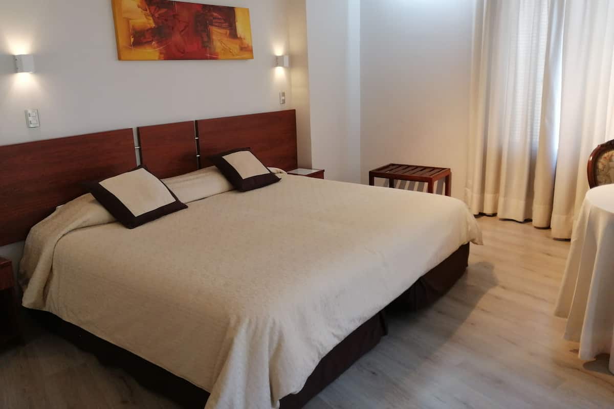 Cama bem arrumada com cabeceira de madeira, mesa de cabeceira e um banquinho de madeira.