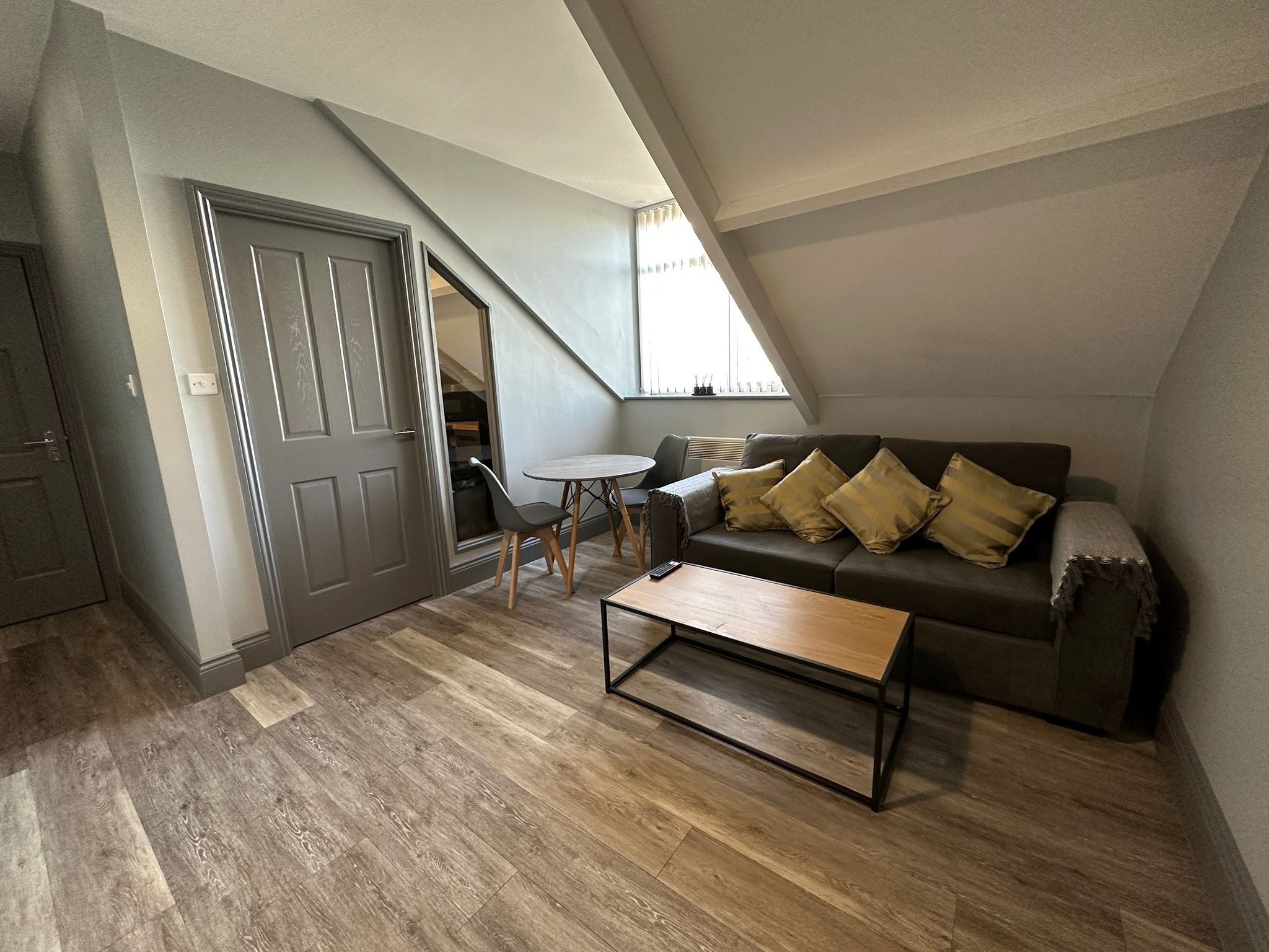 Um quarto estilo loft com teto inclinado, um sofá verde escuro com almofadas, uma mesa de centro de madeira e uma pequena mesa redonda com cadeiras.