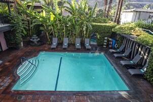 Uma piscina retangular com um deck ladrilhado, rodeada por cadeiras de descanso e uma pequena fonte.