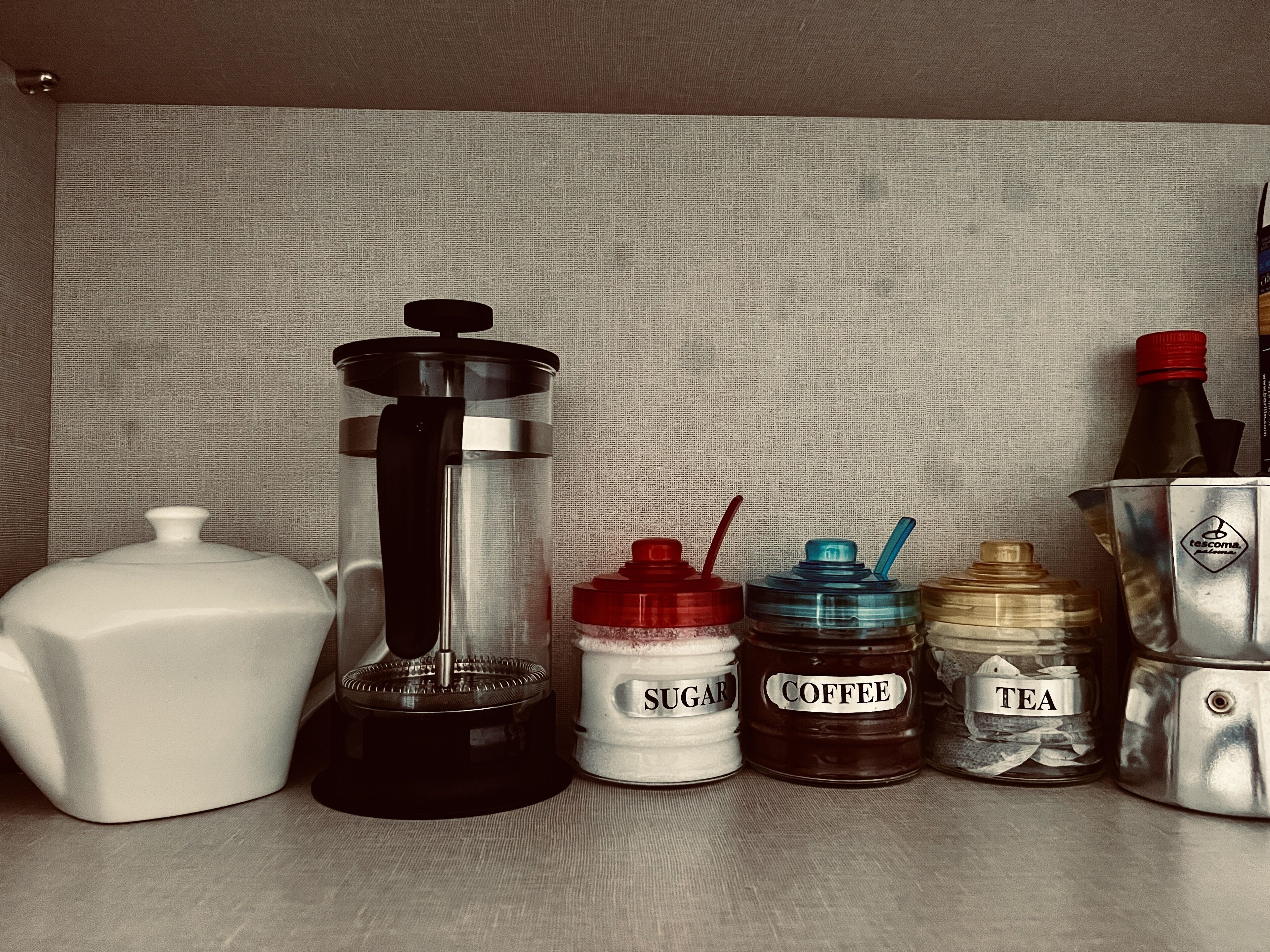 Uma bancada de cozinha com uma prensa francesa, um açucareiro, um recipiente para café e um para chá.