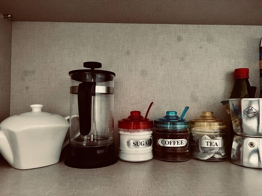 Uma bancada de cozinha com uma prensa francesa, um açucareiro, um recipiente para café e um para chá.