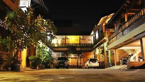 Vista noturna de um edifício com entrada coberta, plantas em vasos e veículos estacionados.