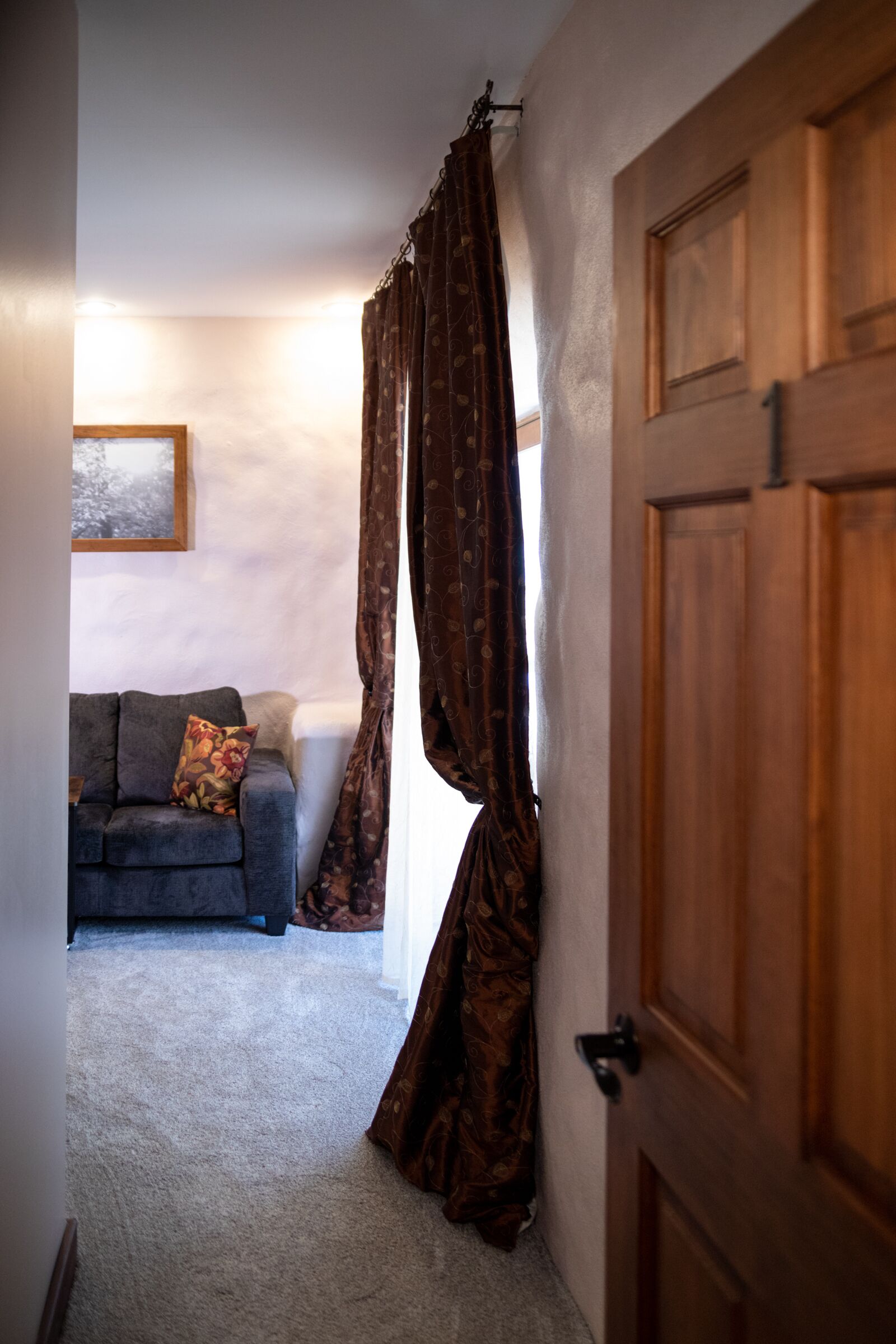 Um corredor com porta de madeira, um sofá cinza com almofada floral e cortinas estampadas.