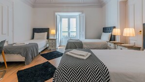 Quarto de hotel com duas camas, uma mesa e uma cadeira. Tem uma janela com vista para uma varanda.