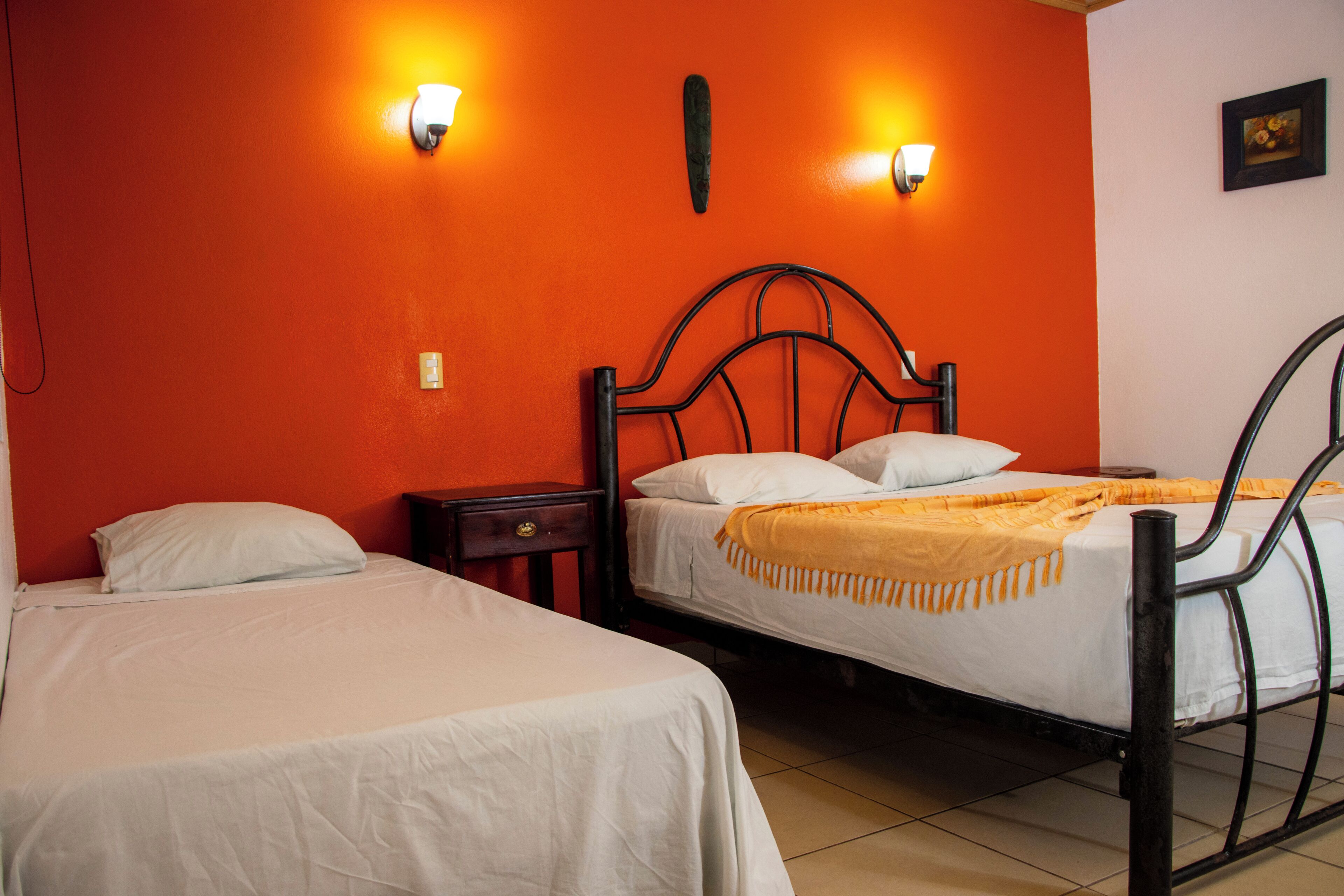 Quarto com duas camas, cada uma com roupa de cama branca e cabeceira de madeira. Há duas luminárias fixadas na parede, sobre um fundo laranja, e uma pequena mesa de cabeceira de madeira com gaveta.