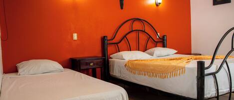 Quarto com duas camas, cada uma com roupa de cama branca e cabeceira de madeira. Há duas luminárias fixadas na parede, sobre um fundo laranja, e uma pequena mesa de cabeceira de madeira com gaveta.