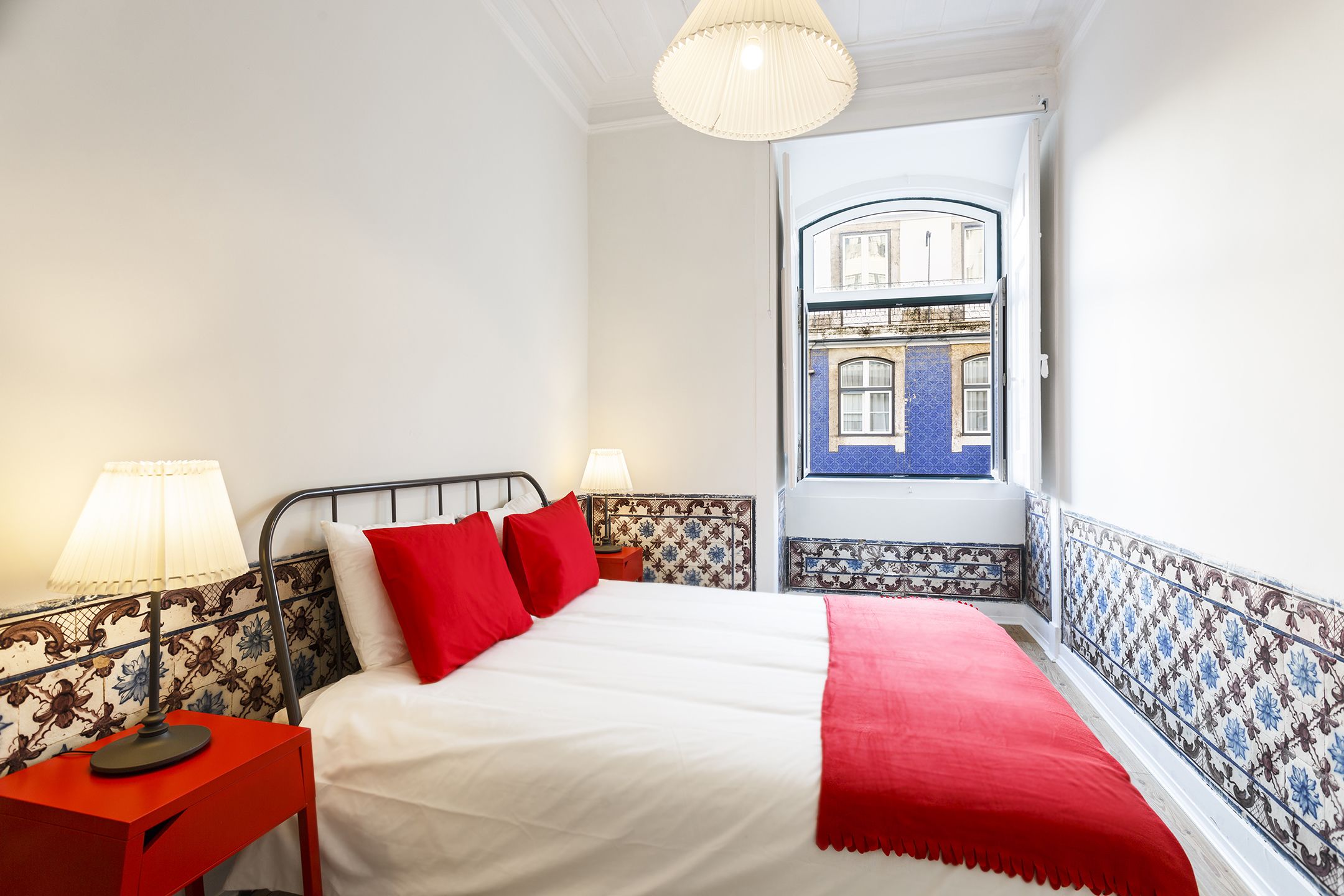 Quarto com cama, duas mesas de cabeceira, janela com vista para um edifício e papel de parede estampado.