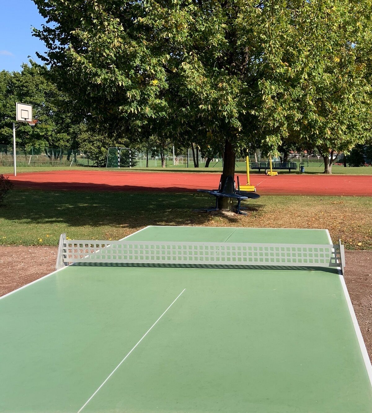 Uma quadra de tênis com rede, rodeada por árvores e uma pista de corrida vermelha.