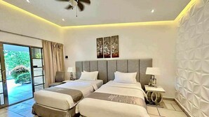 Quarto de hotel moderno com duas camas, ventilador de teto, janela grande com cortinas e vista para o verde lá fora.