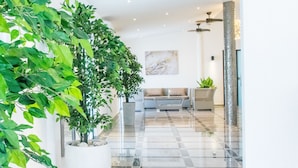 Um corredor moderno com plantas em vasos, área de estar com sofá e cadeiras, e um ventilador de teto.