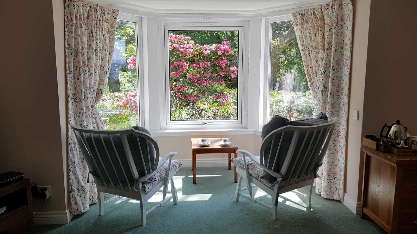 Quarto com duas cadeiras viradas para uma mesa, uma janela com cortinas florais e vista para flores cor-de-rosa lá fora.