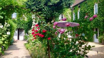 Um caminho de jardim com pérgola coberta por plantas trepadeiras e flores, conduzindo a um edifício com fachada branca e janelas.