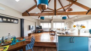 Cozinha moderna com vigas de madeira, área de refeições e sala de estar com sofá e televisão.