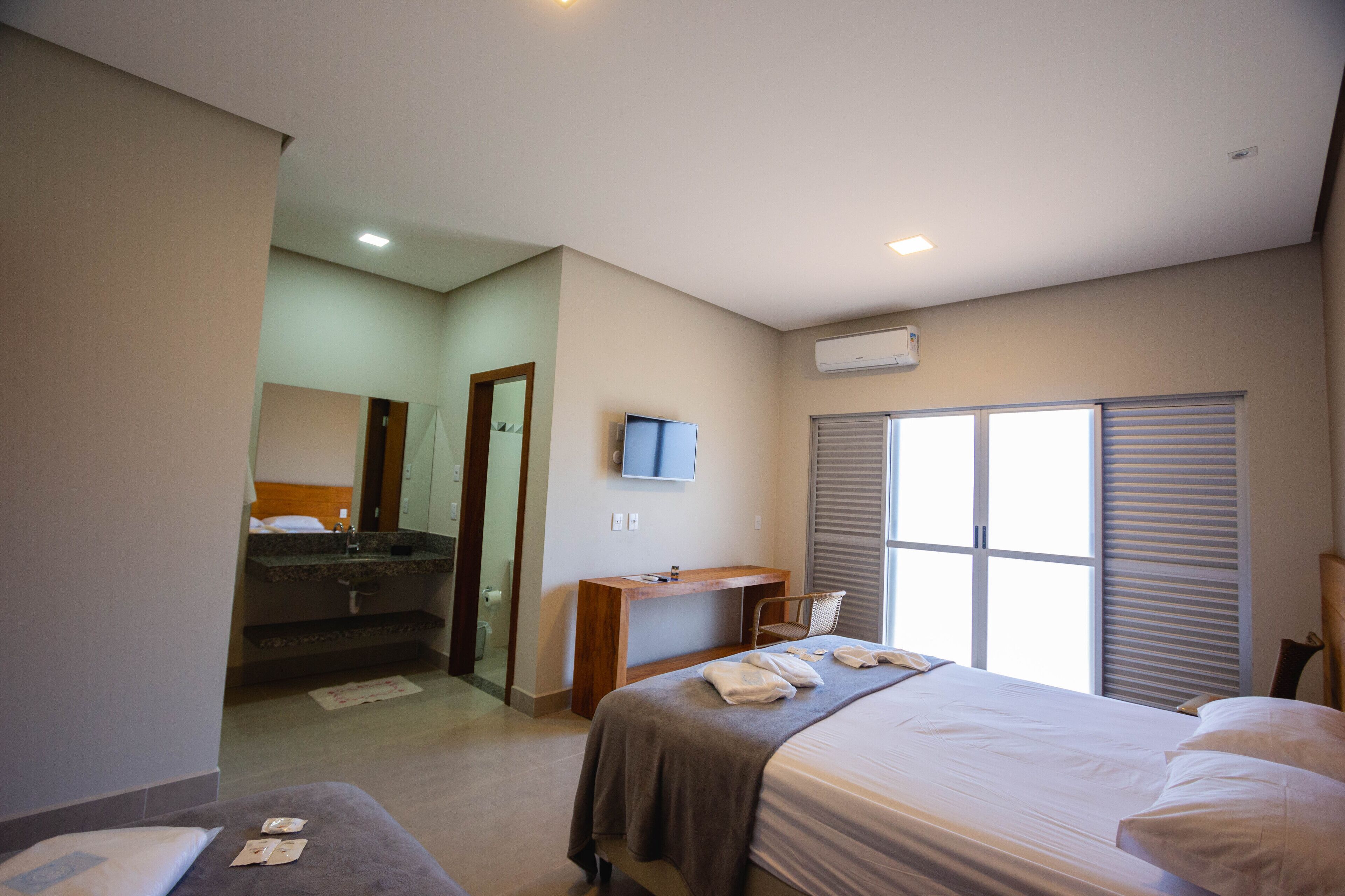 Quarto de hotel com cama, escrivaninha, televisão, janela com persianas e banheiro com pia e espelho.