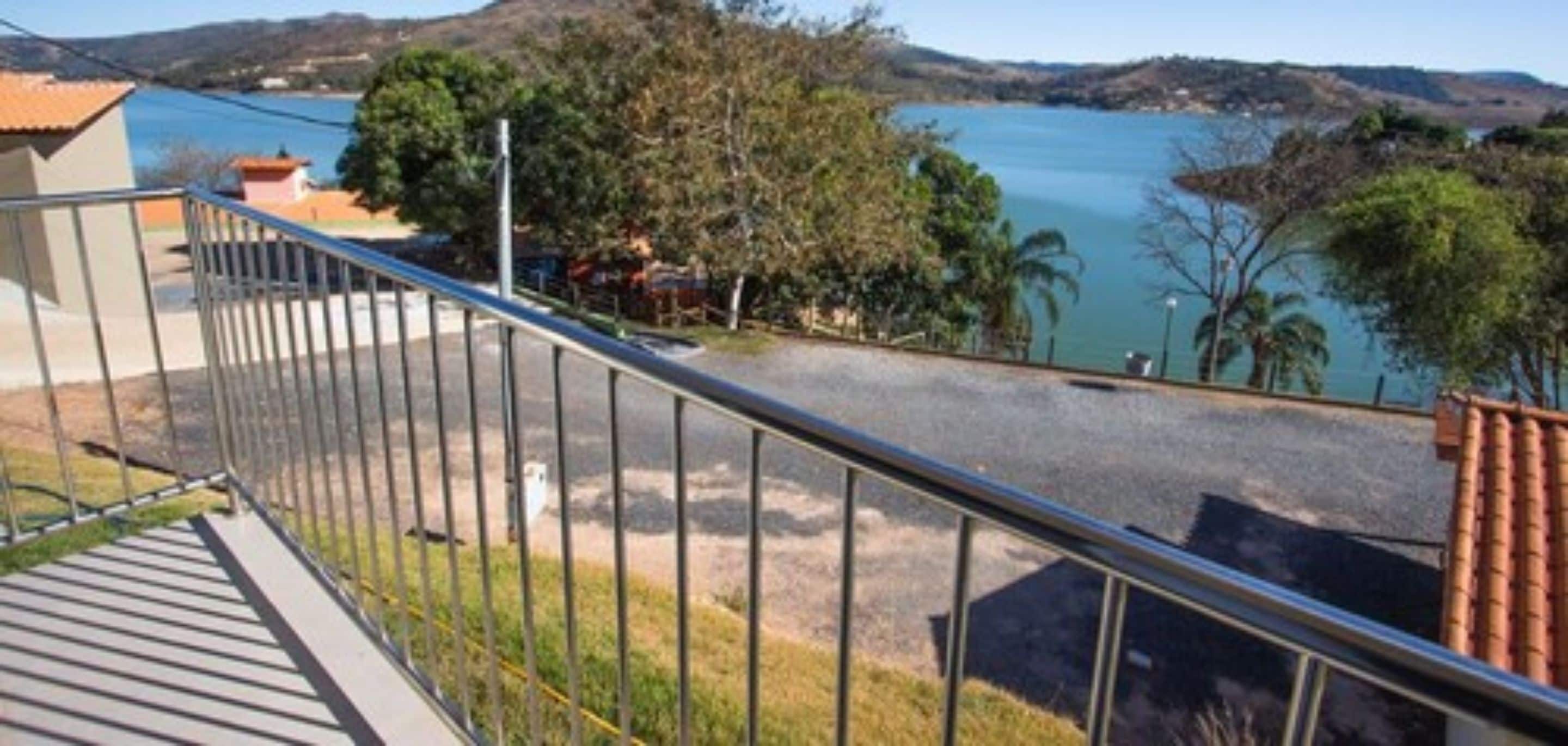 Varanda com grade de metal dando para um lago, cercada por árvores e uma paisagem montanhosa.
