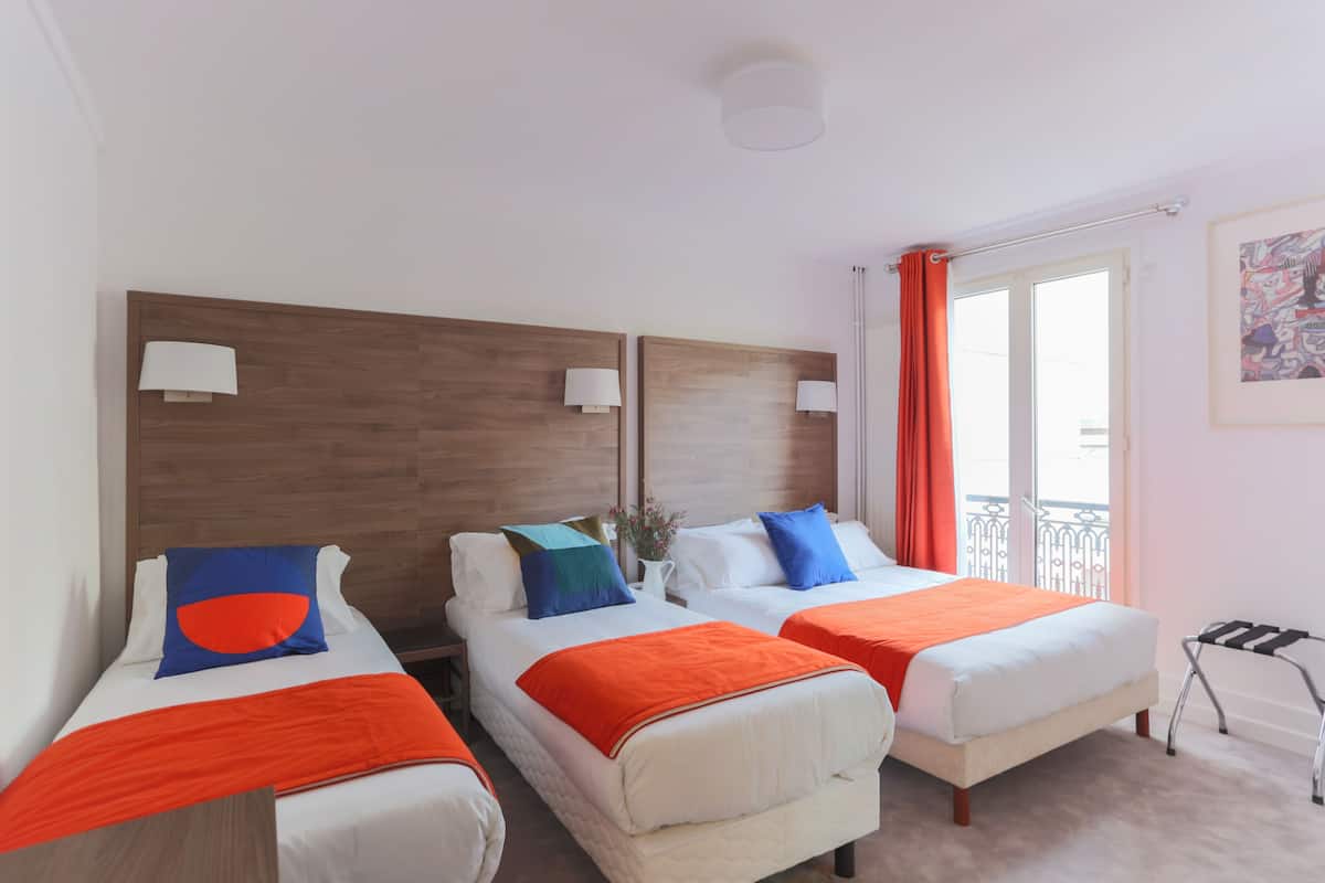 Quarto de hotel com duas camas, cada uma com roupa de cama branca e mantas laranja. Há duas mesas de cabeceira com abajures, uma janela com cortinas laranja e um quadro na parede.