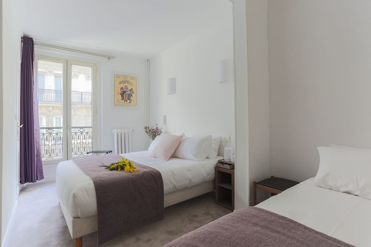 Quarto de hotel com cama, mesa de cabeceira, janela com cortinas, radiador e um pôster na parede.
