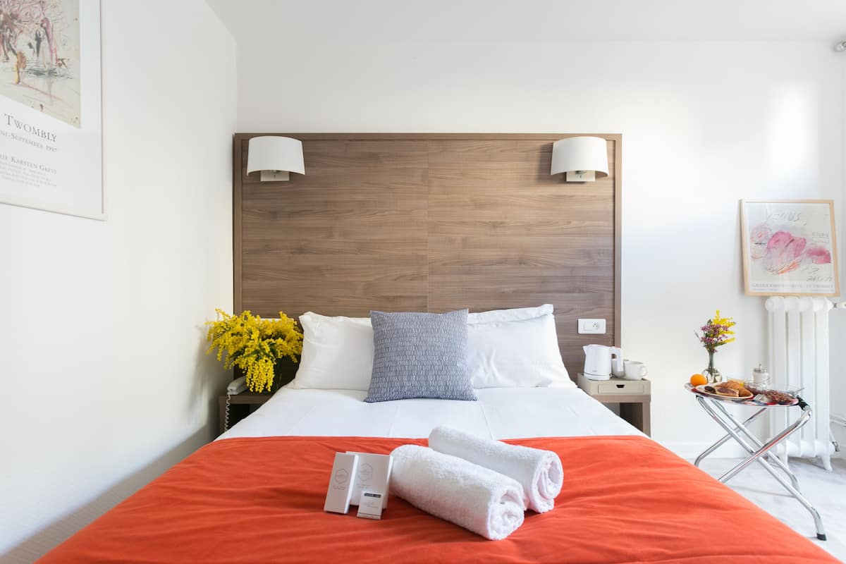Cama bem arrumada com colcha vermelha, travesseiros brancos e um travesseiro azul. Há duas luminárias brancas de cada lado da cabeceira, uma mesinha com uma bandeja de comida e um vaso de flores, e um quadro na parede.