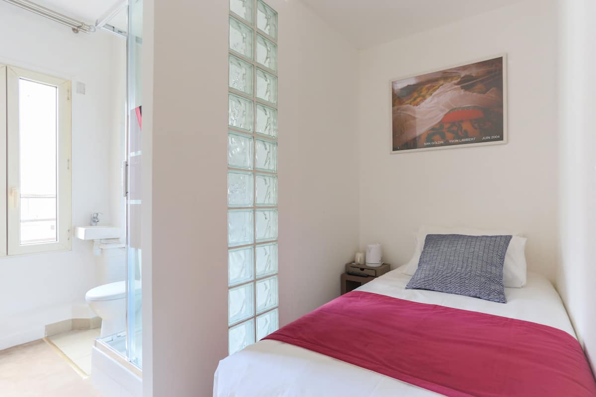 Quarto com cama, mesa de cabeceira, janela com vidro jateado e um quadro na parede.