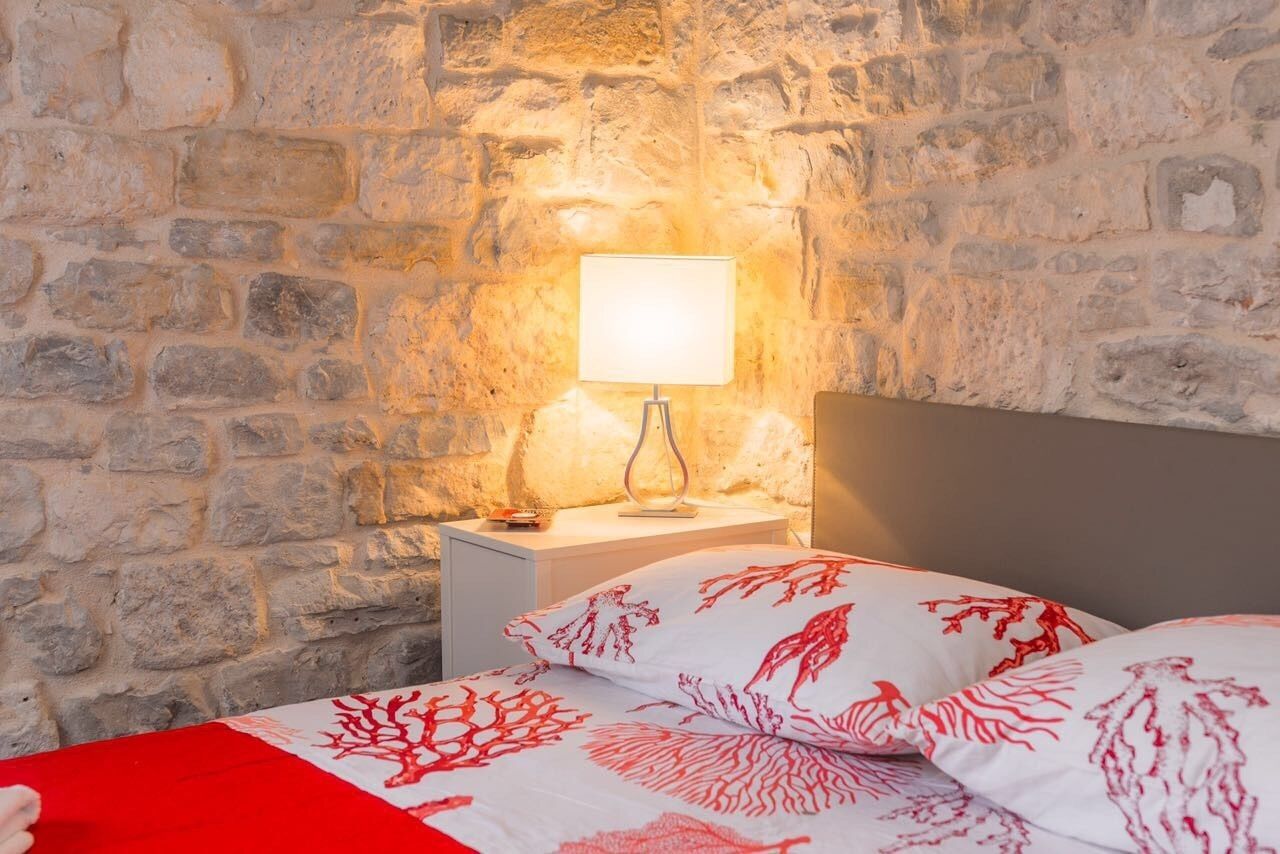 Quarto com parede de pedra, mesa de cabeceira com abajur e cama com roupa de cama vermelha e branca.