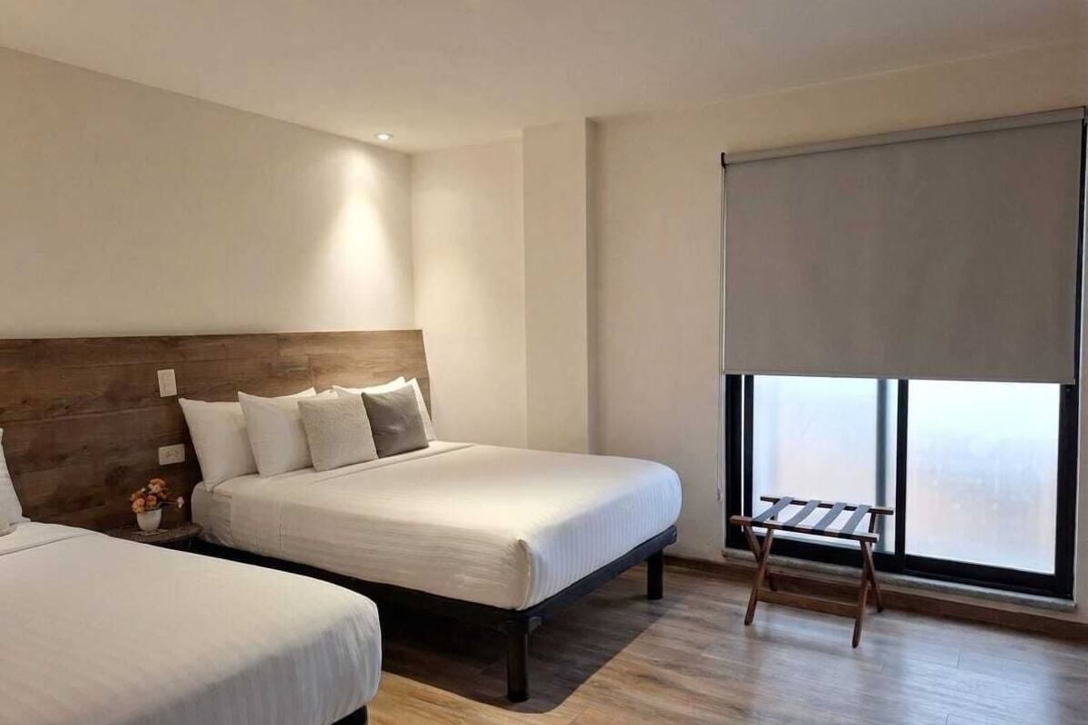 Quarto de hotel com duas camas, uma cabeceira de madeira, uma mesinha e uma janela com vista.