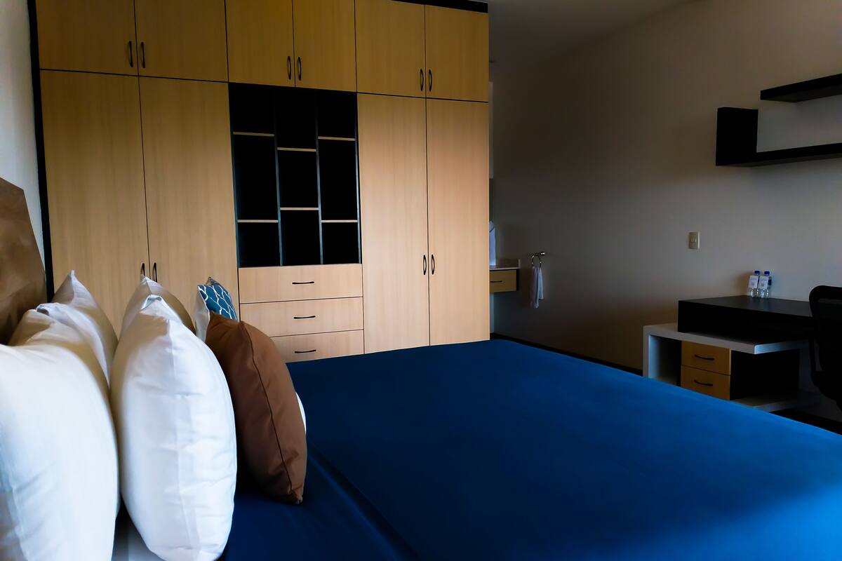 Quarto com uma cama grande, guarda-roupa com vários compartimentos, escrivaninha com cadeira e uma pequena mesa com toalha.