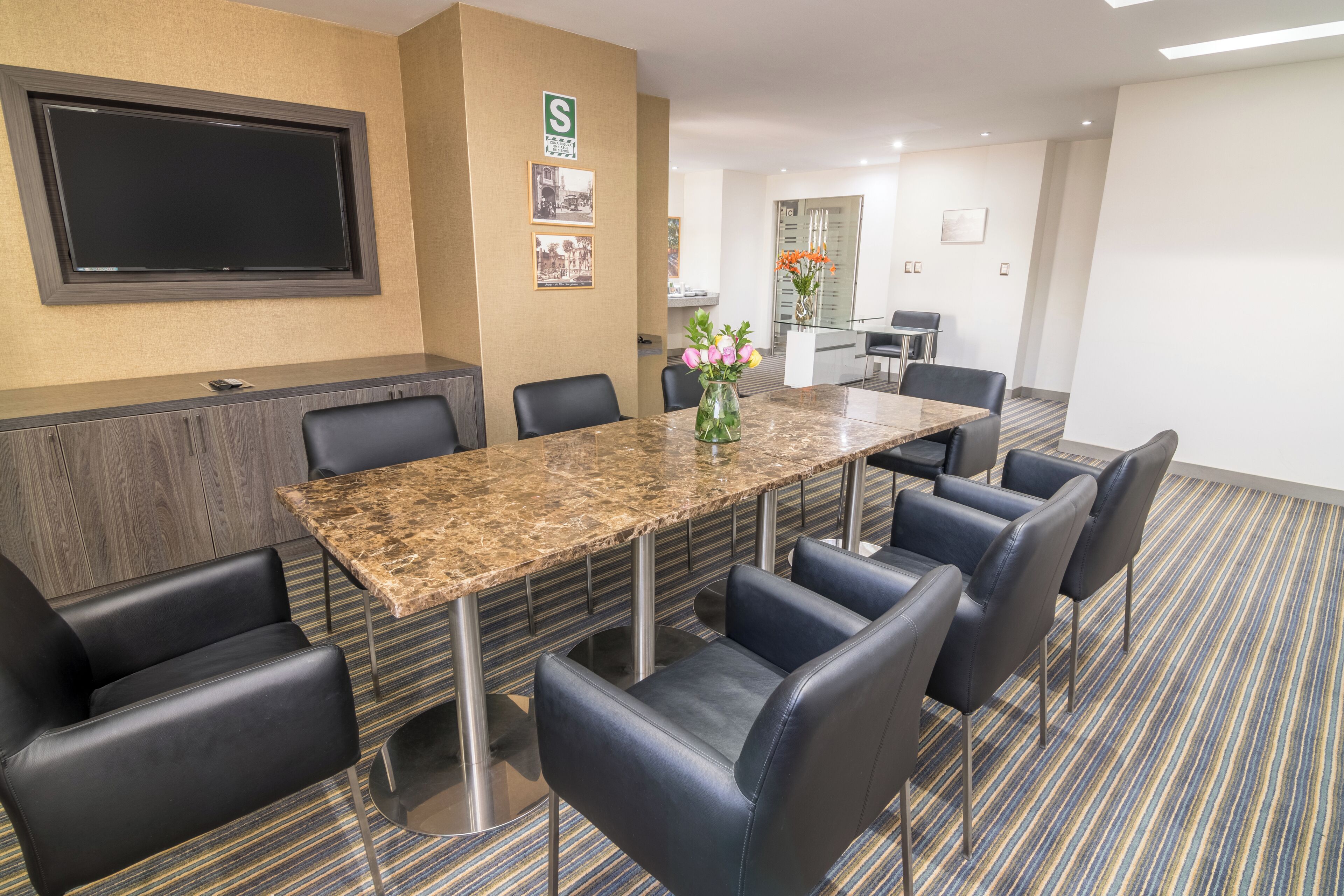 Sala de conferência com mesa de mármore, cadeiras de couro preto, TV fixada na parede e piso coberto de carpete.