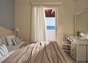 Quarto com cama, mesa de cabeceira, cômoda e vista para o mar através de uma porta.