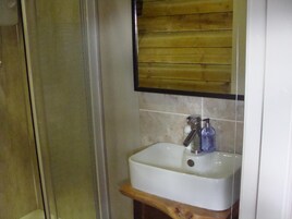 Casa de banho com cabine de chuveiro em vidro, pia branca com prateleira de madeira e espelho com moldura de madeira.